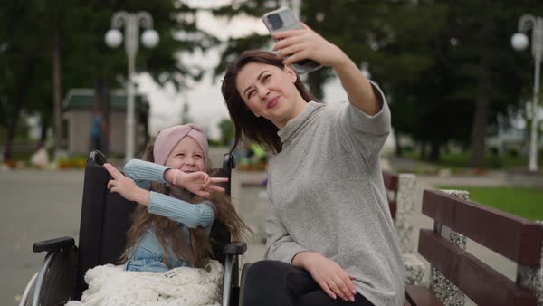 Mamma takes selfies while little girl with disability making funny gestures with hands. Woman ...