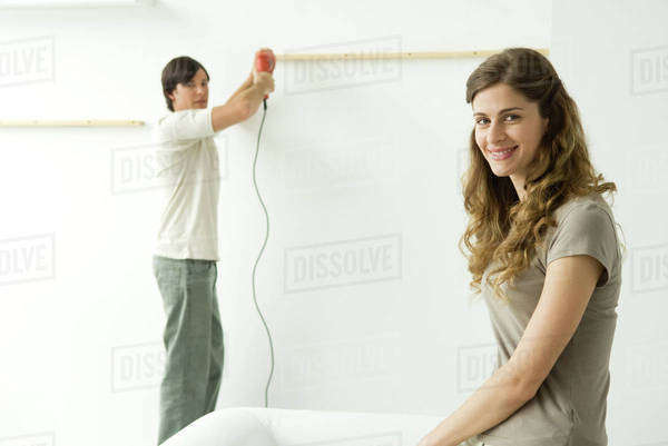 Man Attaching Thin Piece Of Wood To Wall Woman In Foreground Smiling At Camera Stock Photo man-attaching-thin-piece-of-wood-to-wall-woman-in-foreground-smiling-at-camera-stock-photo