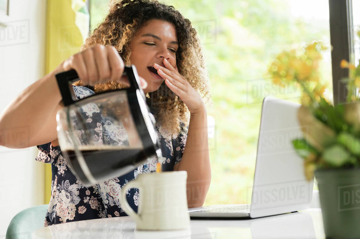 Tired woman pouring coffee by laptop - Royalty-free Stock Photo | Dissolve