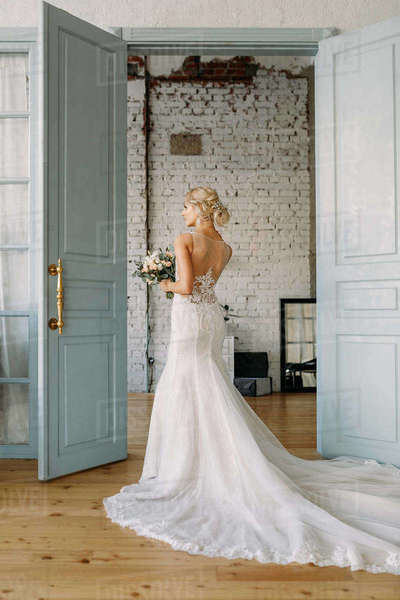 The blonde bride stands half-turned at the open door in a wedding dress ...