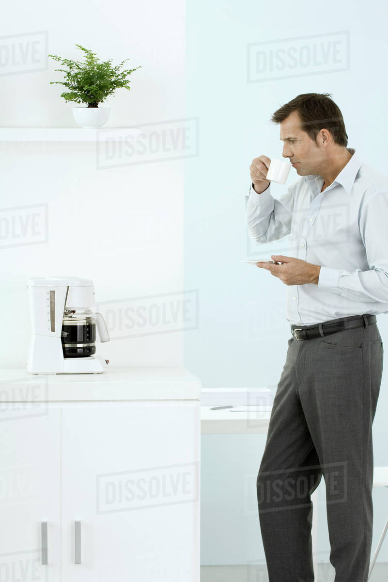 Man standing by coffee maker, taking a sip of coffee, holding saucer ...