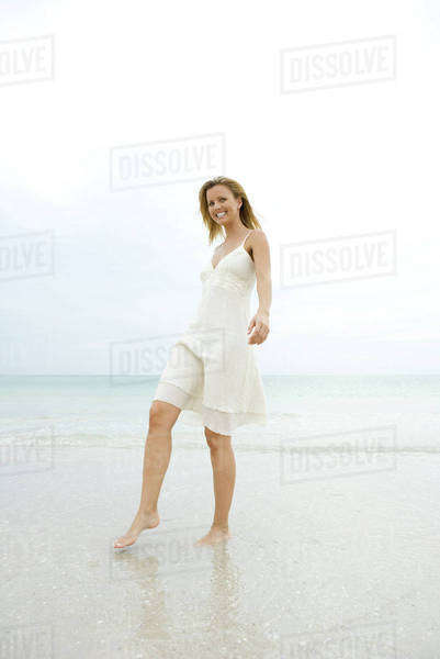 Young woman in sundress walking on wet sand at beach, smiling at camera ...