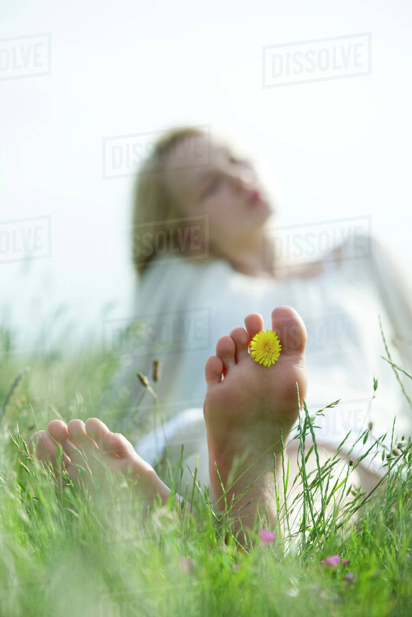 Barefoot young woman sitting in grass, holding dandelion flower between ...