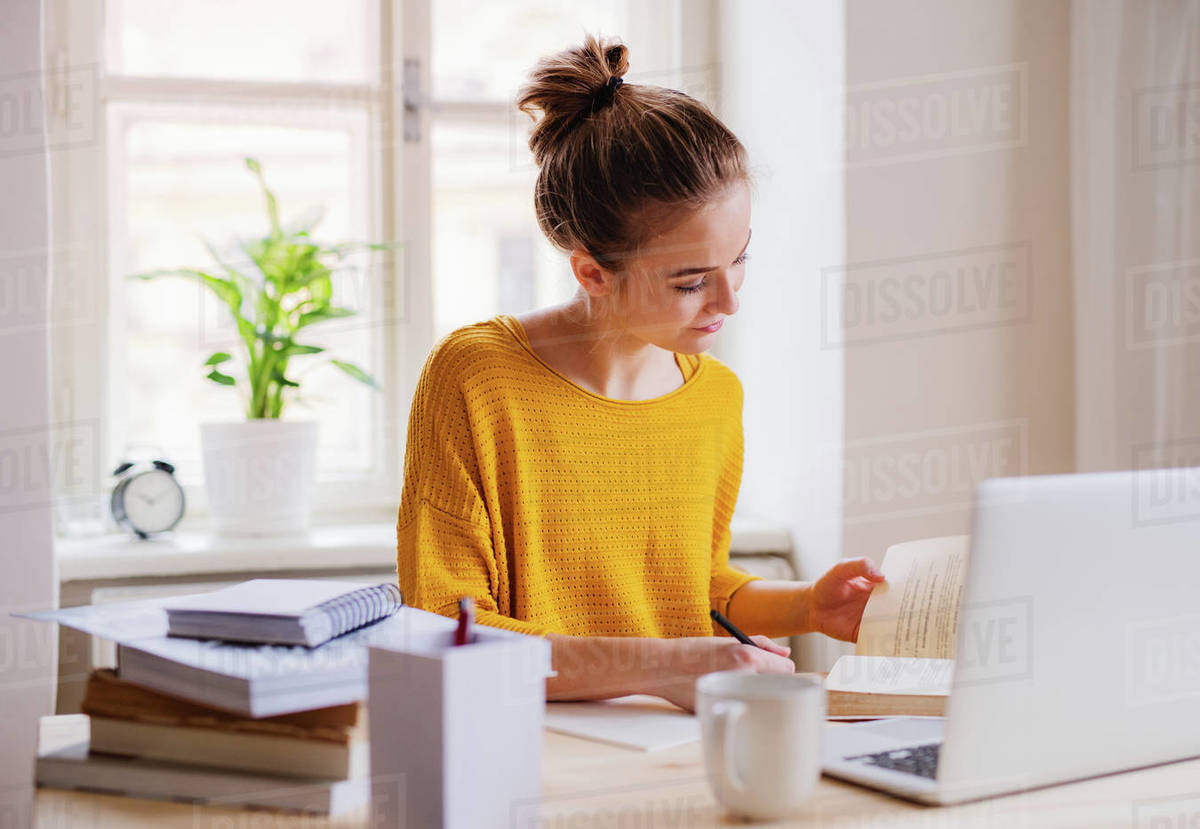 A young happy college female student sitting at the table at home ...