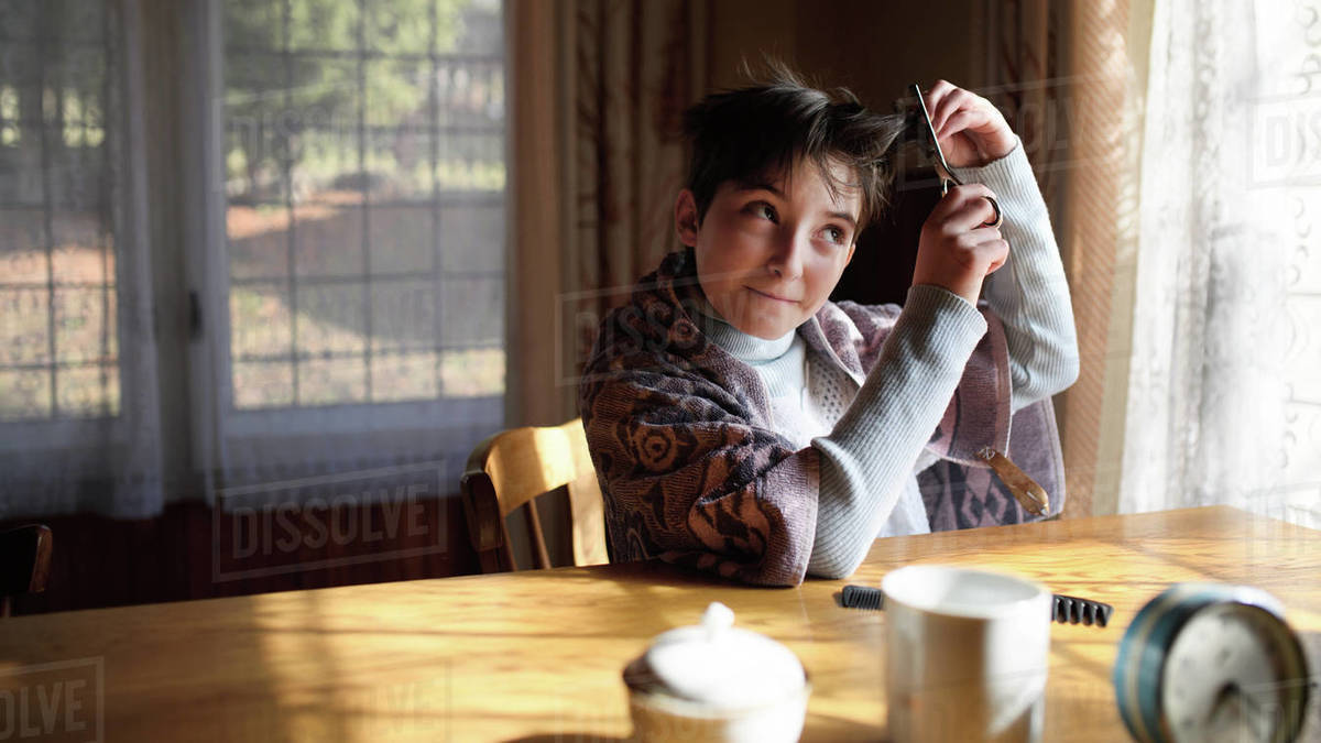 Poor sad small girl sitting at the table and cutting hair indoors at ...