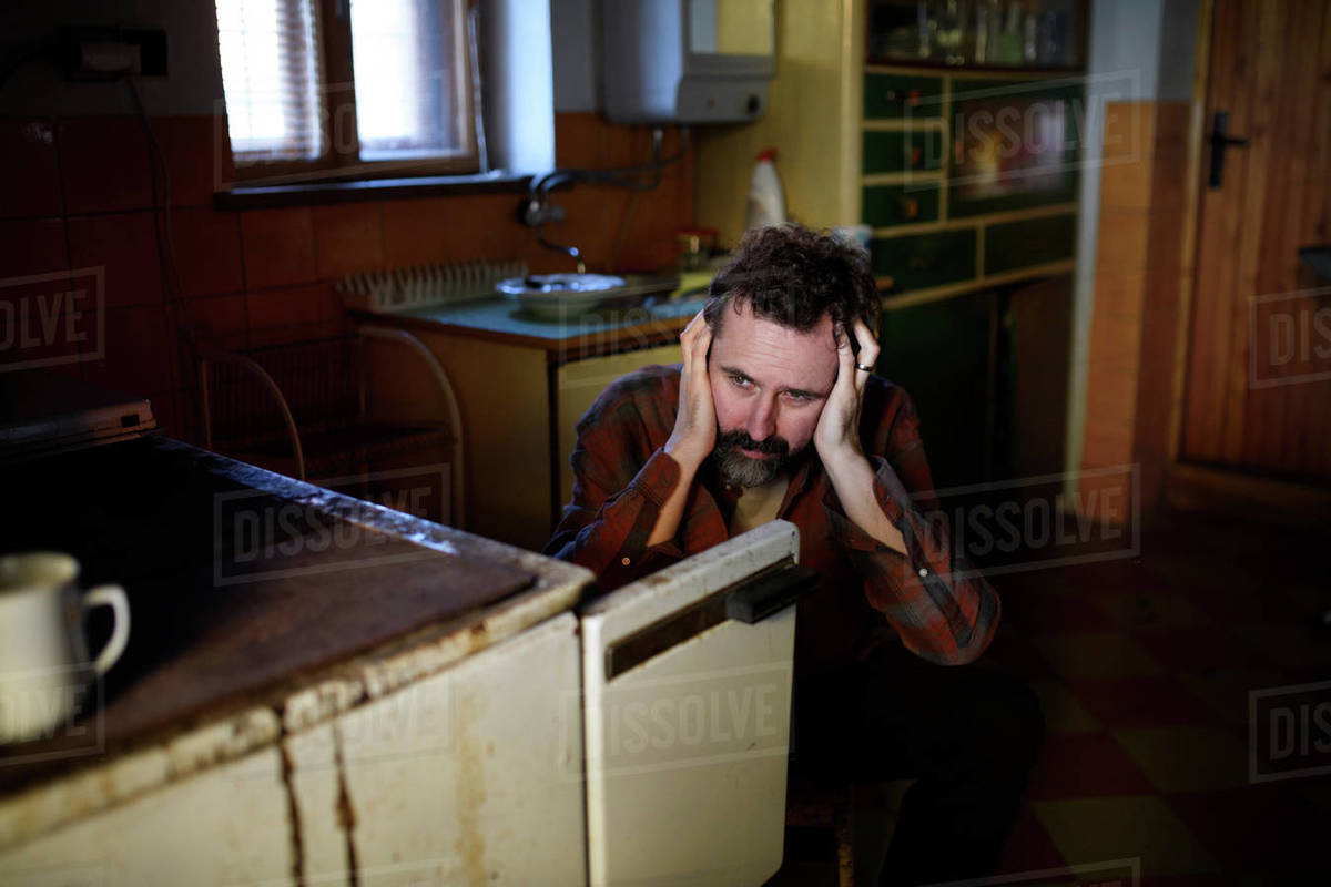A portrait of sad poor mature man sitting indoors at home, poverty ...