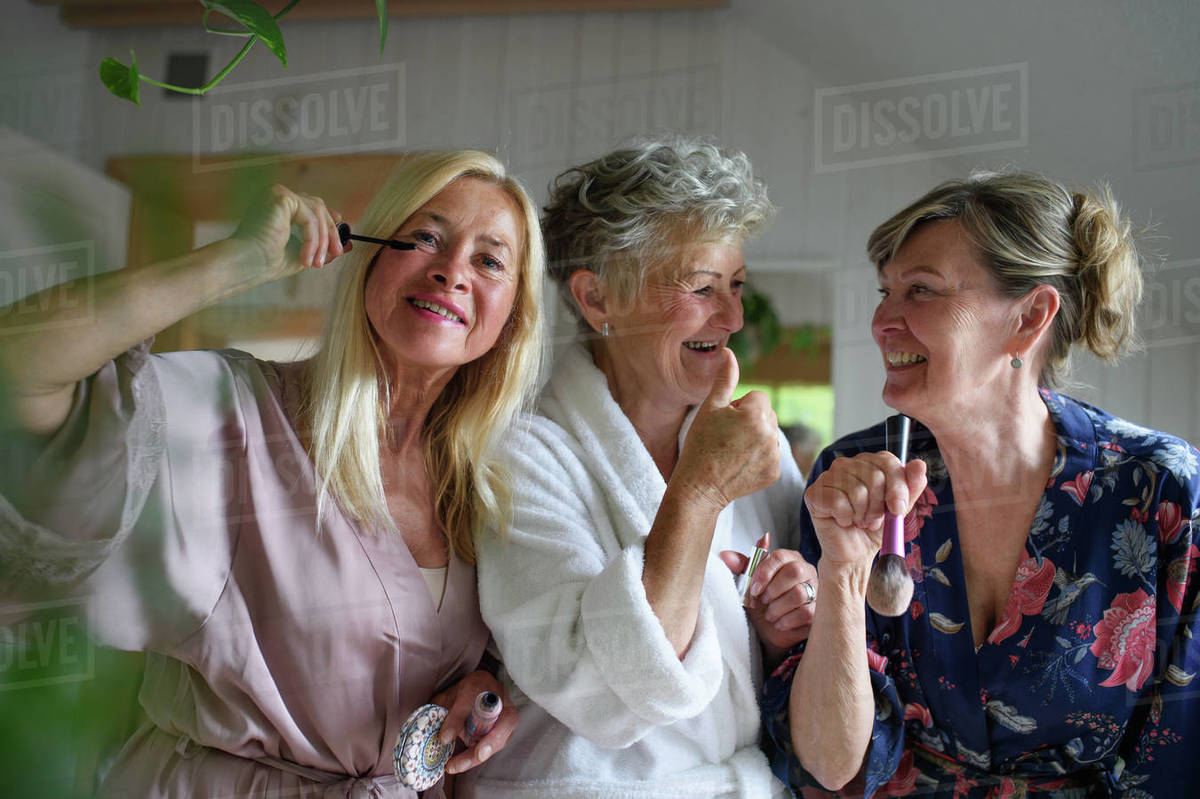 Happy senior women friends in bathrobes doing a make up indoors at home ...