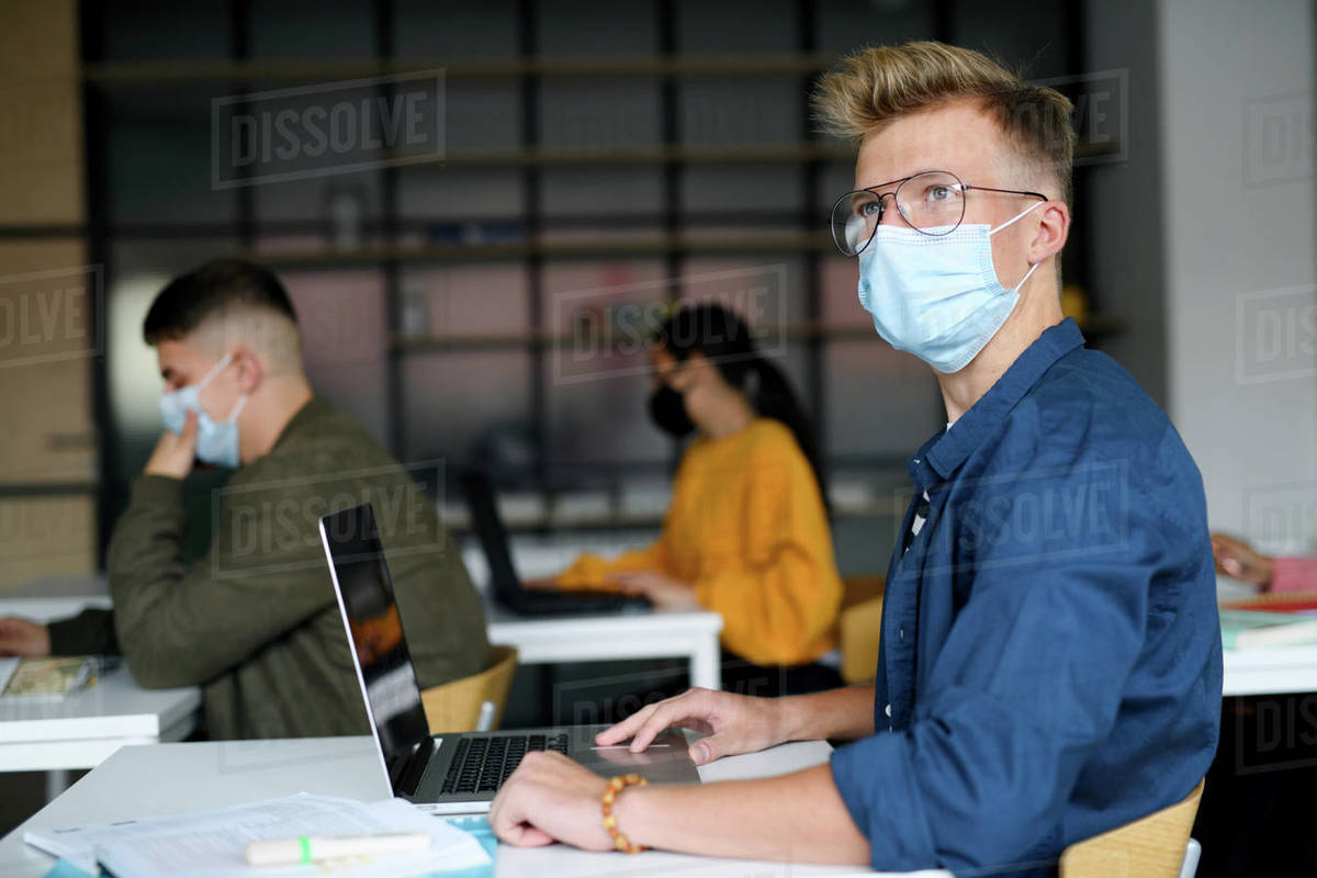 Side view of young students with face masks at desks at college or ...