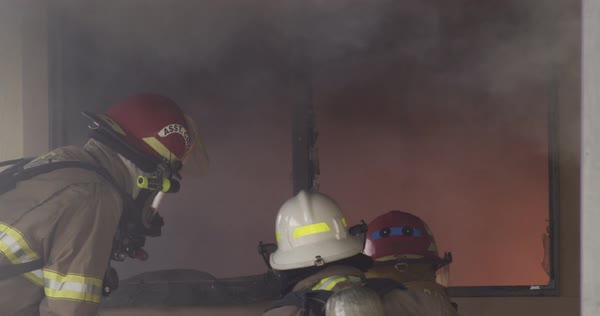Three firefighters in front of a glowing window spray a stream of water ...