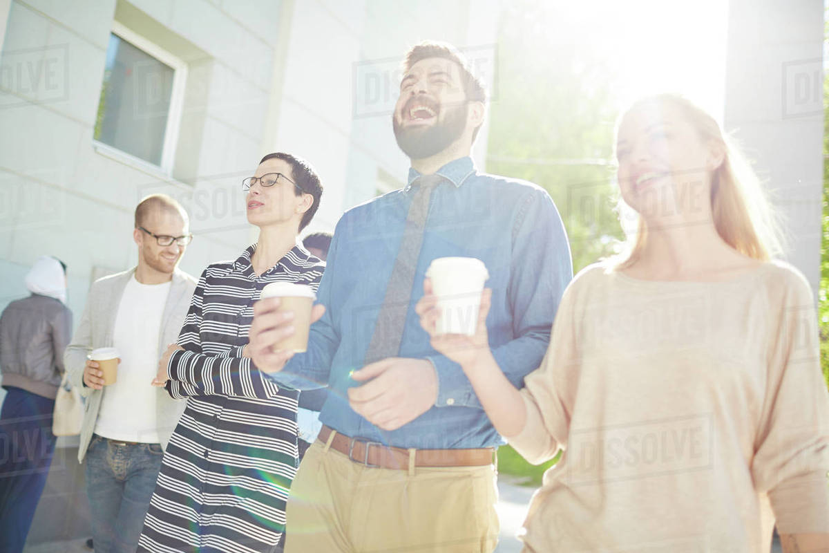 Group of joyful colleagues with drinks having walk and enjoying sunny ...