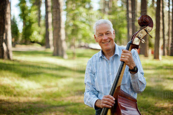 Portrait of a smiling senior man playing a double bass in a forest ...