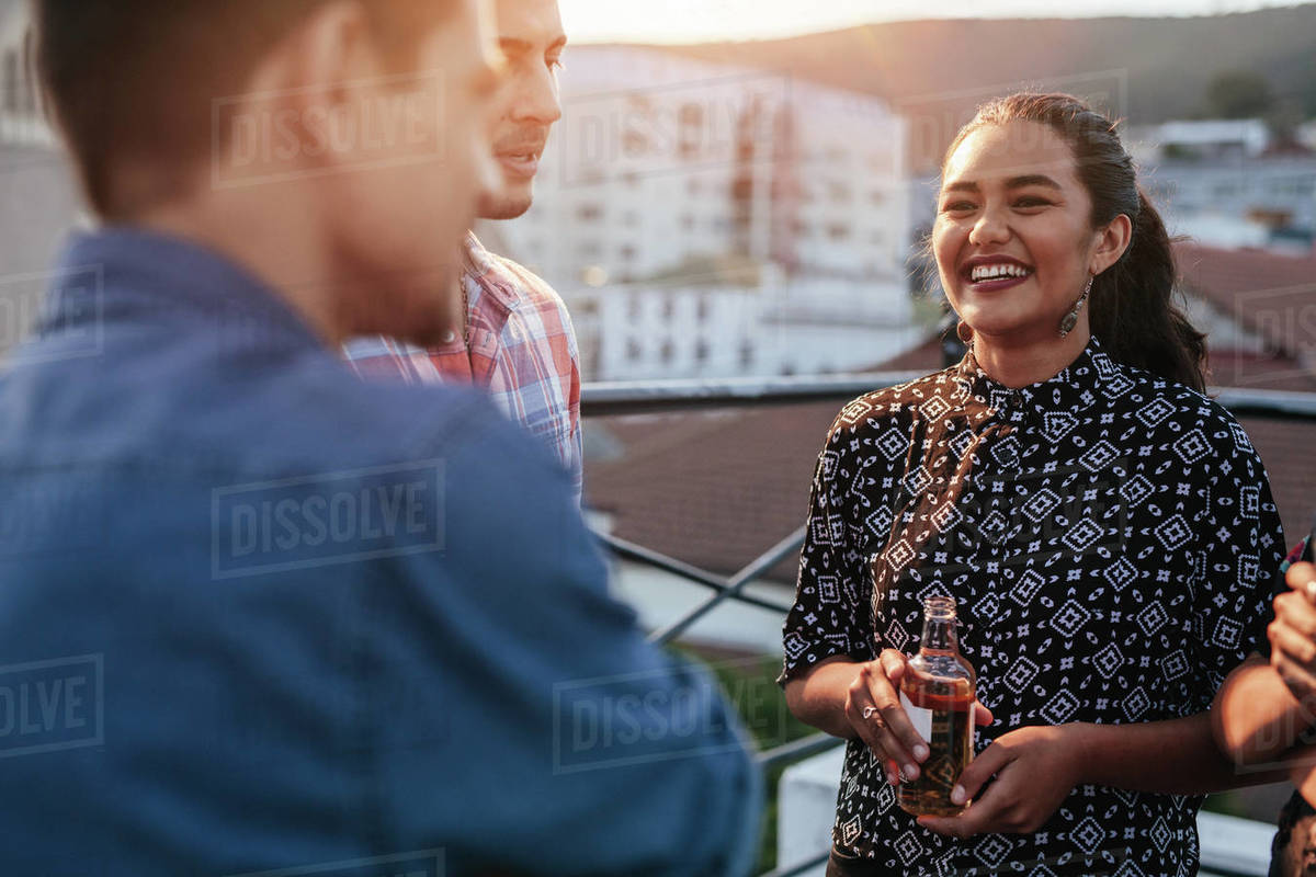 Smiling young woman holding beer standing on rooftop with friends ...