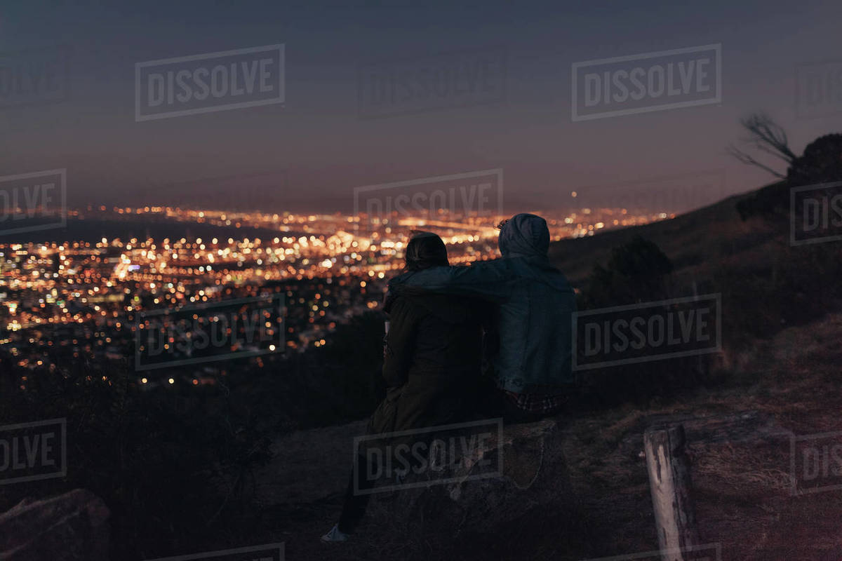 Couple watching the brightly lit city sitting on top of a hill late in ...