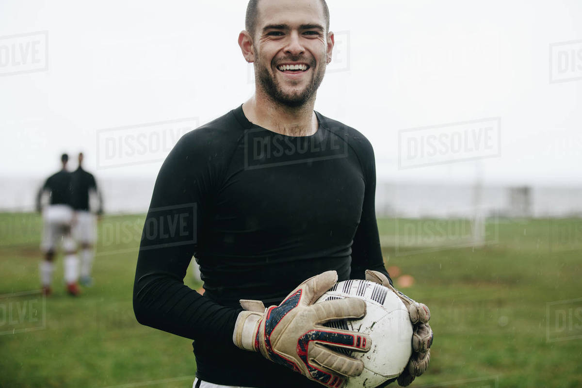 Portrait of a cheerful goalkeeper standing with a soccer ball in hand ...