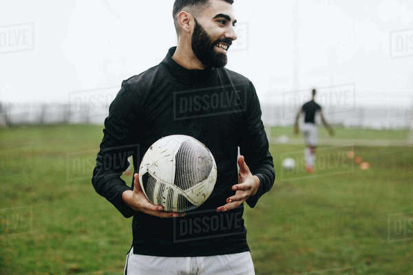 Smiling footballer standing on field holding a football during practice ...