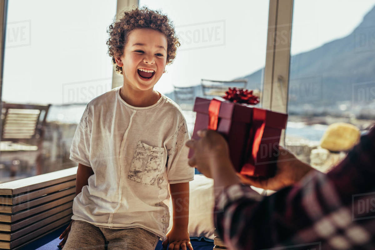 Person giving a gift box to a boy sitting in a beach house. Excited kid ...