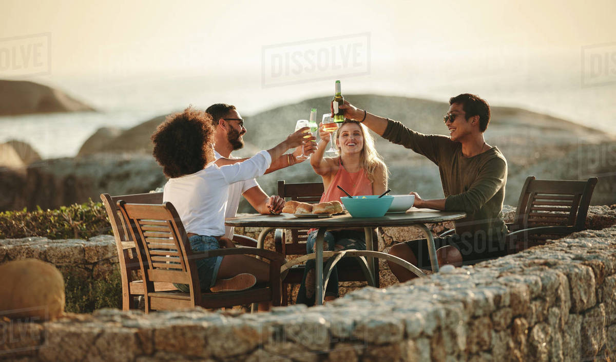 Four friends toasting drinks sitting on a dining table outdoors near ...