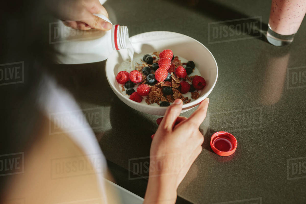 Woman adding milk in bowl with berries and cereals on kitchen counter ...