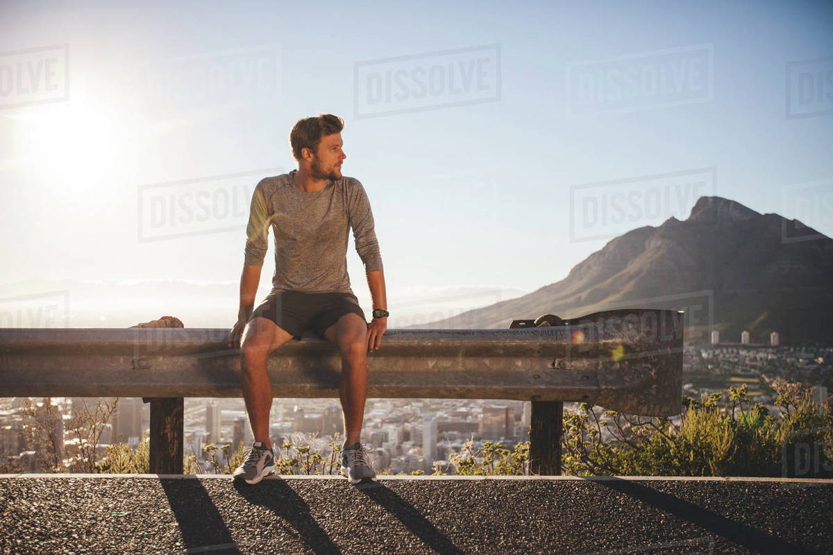 Male runner sitting on a guardrail on country road looking away on ...