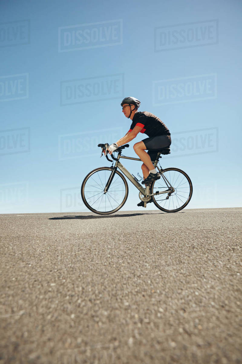 Side view of male cyclist riding bicycle on flat road against blue sky ...