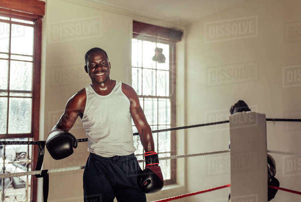 Happy sweaty fit young African boxer leaning back against the ropes in ...