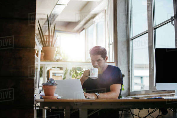 Relaxed young man sitting at his desk working on laptop and drinking ...