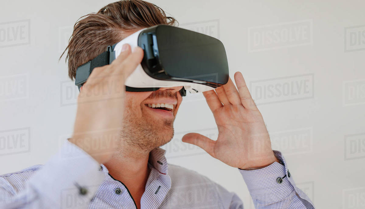 Shot of happy young man wearing virtual reality goggle against grey ...