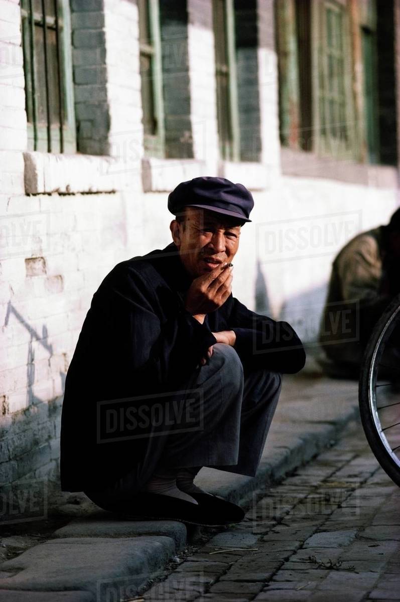 Man smoking a cigarette while squatting on the sidewalk, China ...