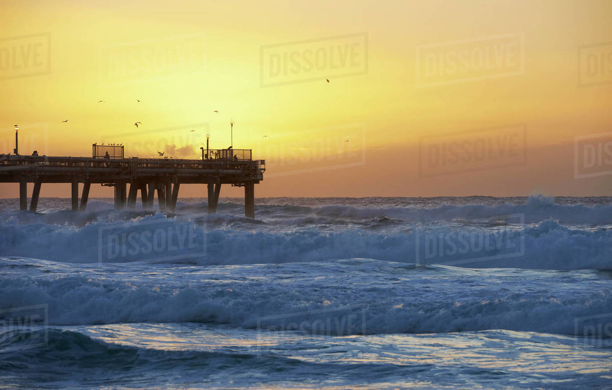 People walking and fishing on ocean pier as the sun set over the ocean ...