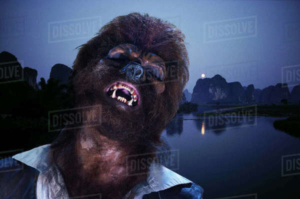 Wolfman howling at the moon standing on rocks in front of lake at night ...