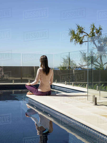 Back view of woman sitting cross legged next to swimming pool ...