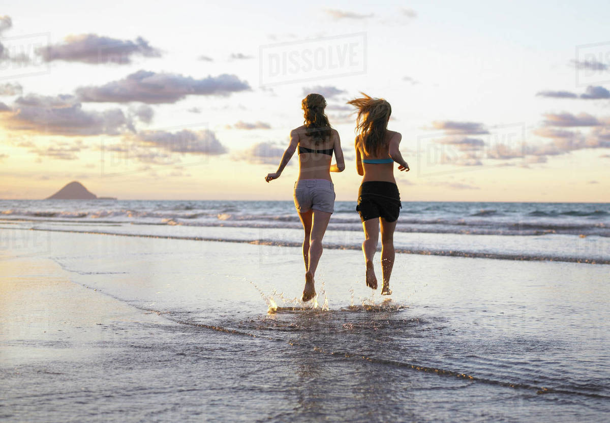 Back view of two young women running in the water along beach in the ...