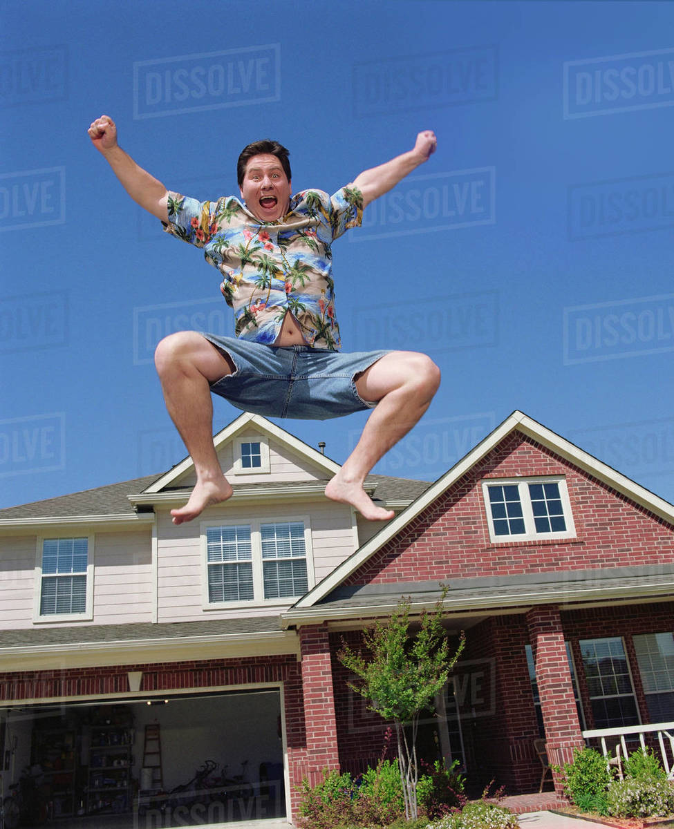 Middle aged man jumping in front of home, striking a pose mid air ...