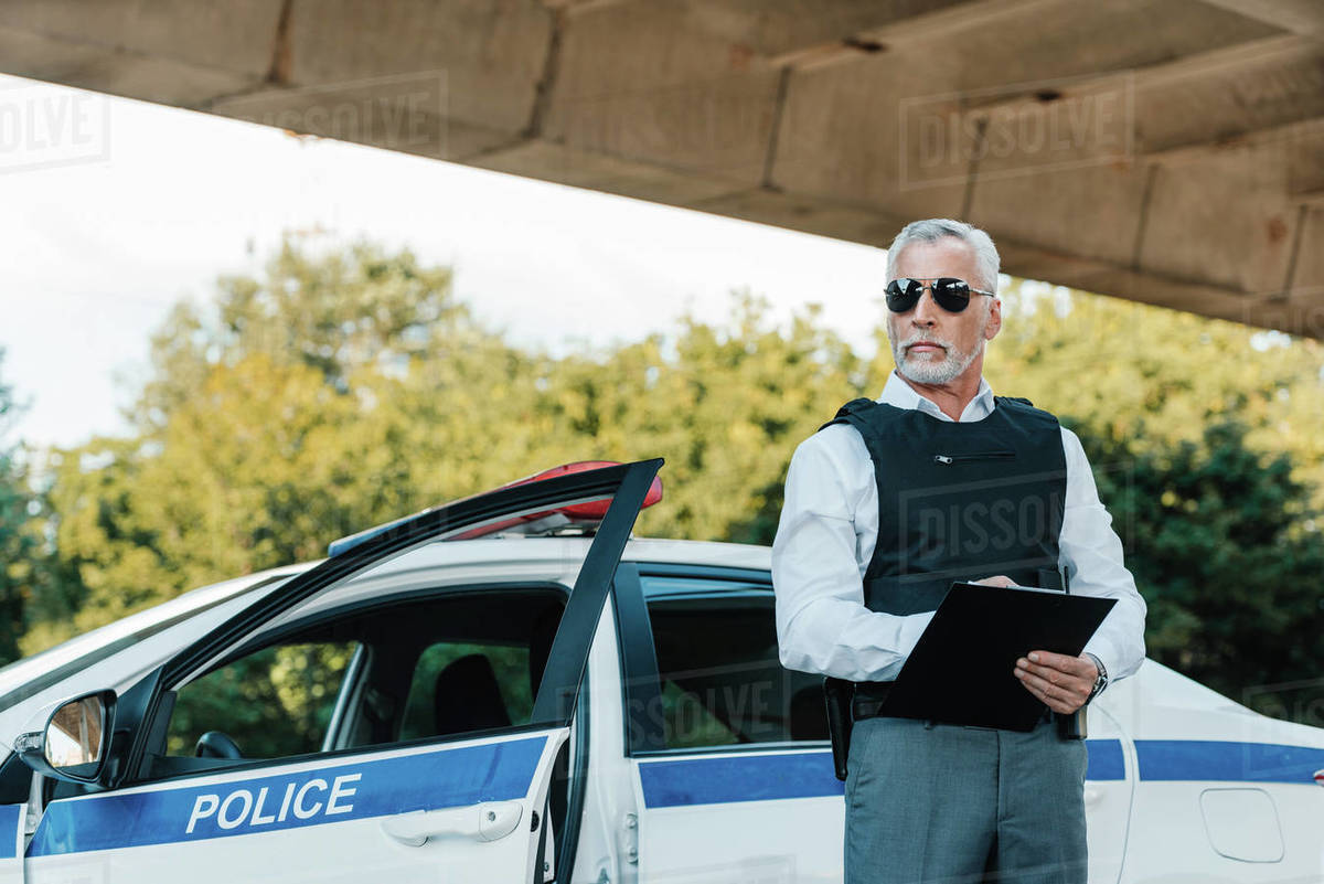 policeman in bulletproof vest and sunglasses writing in clipboard near ...