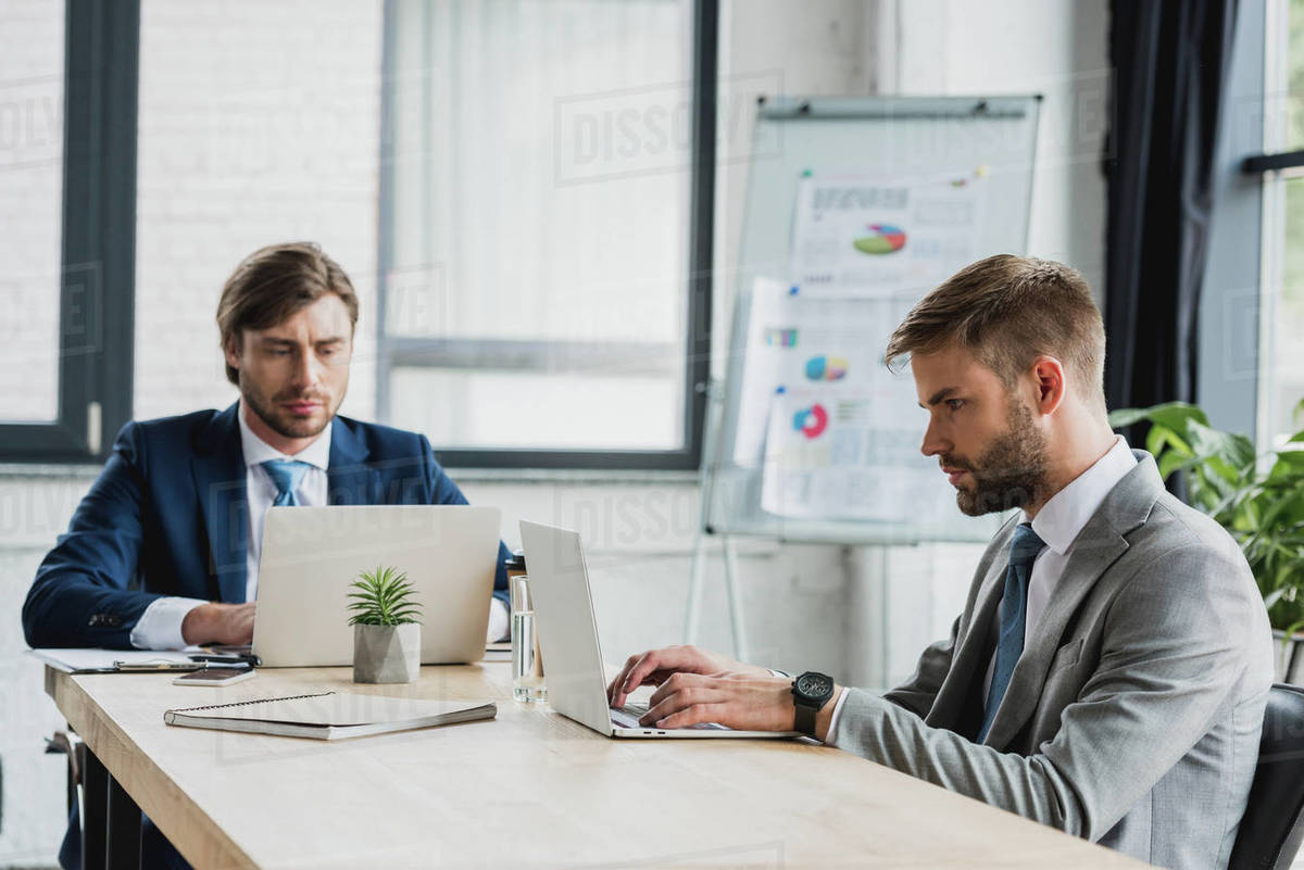 Focused young businessmen in suits using laptops and working together ...