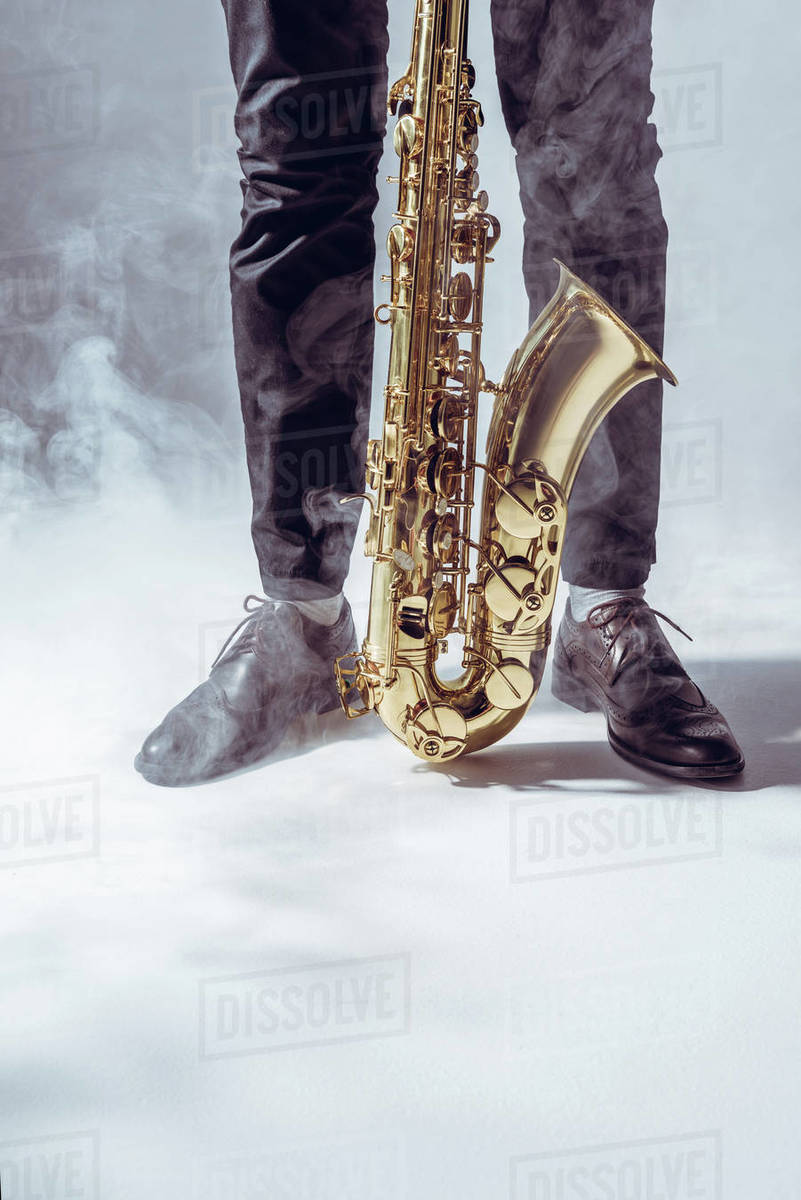 Partial view of young musician standing with saxophone in smoke on grey ...