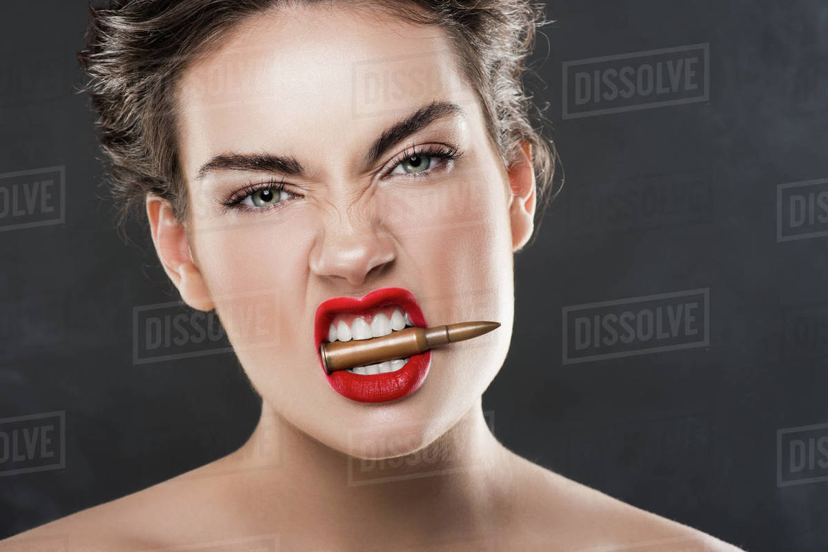 Aggressive elegant woman holding bullet in teeth, isolated on grey ...