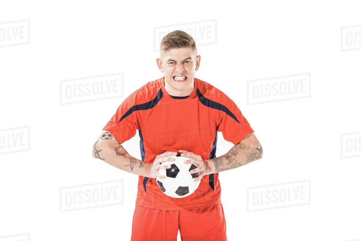 angry young soccer player holding ball and looking at camera isolated ...