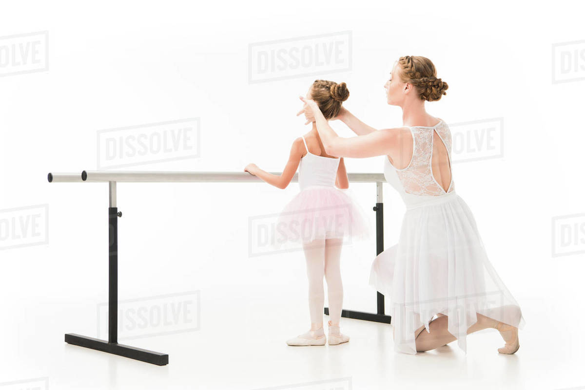 Rear view of adult female teacher in tutu helping little ballerina ...