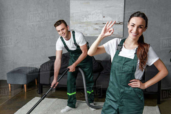 Happy young cleaning company workers using vacuum cleaner and showing ...