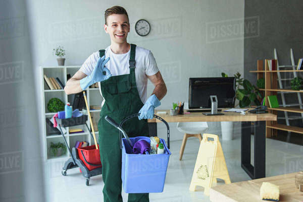Handsome smiling young cleaner holding bucket with cleaning supplies ...
