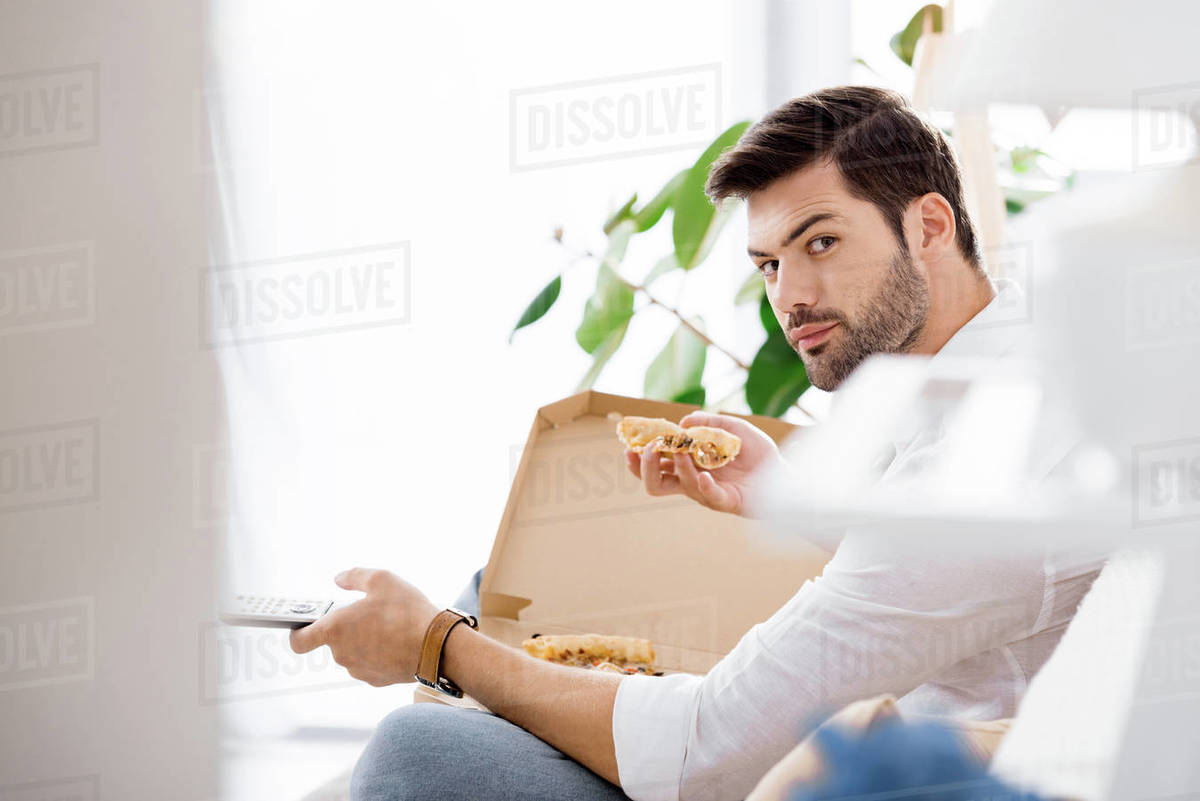 Side view of young man with remote control in hand eating pizza alone ...