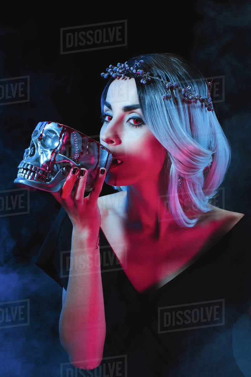 Sexy vampire woman drinking blood from metal skull on dark background ...