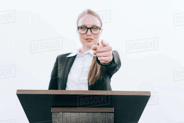 Selective focus of attractive lecturer pointing on camera from podium ...