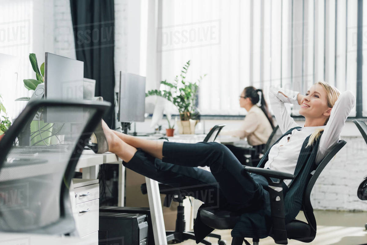 Smiling young businesswoman resting with legs on desk in office ...