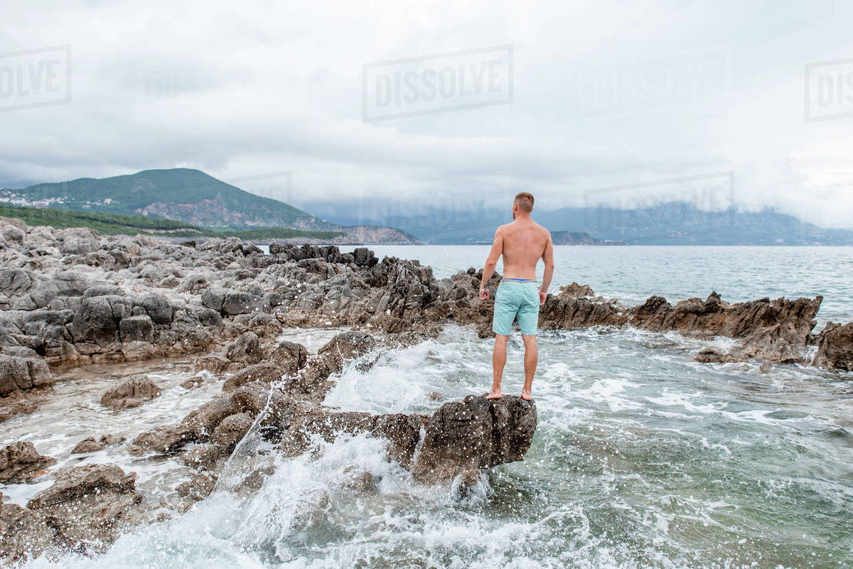 Back view of handsome muscular young man standing on rocky beach and ...