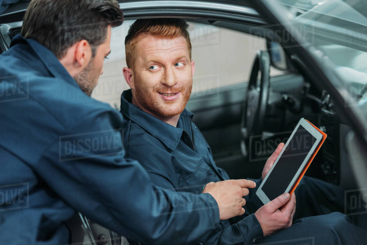 Two car mechanics sitting in a car at workshop using a digital tablet ...