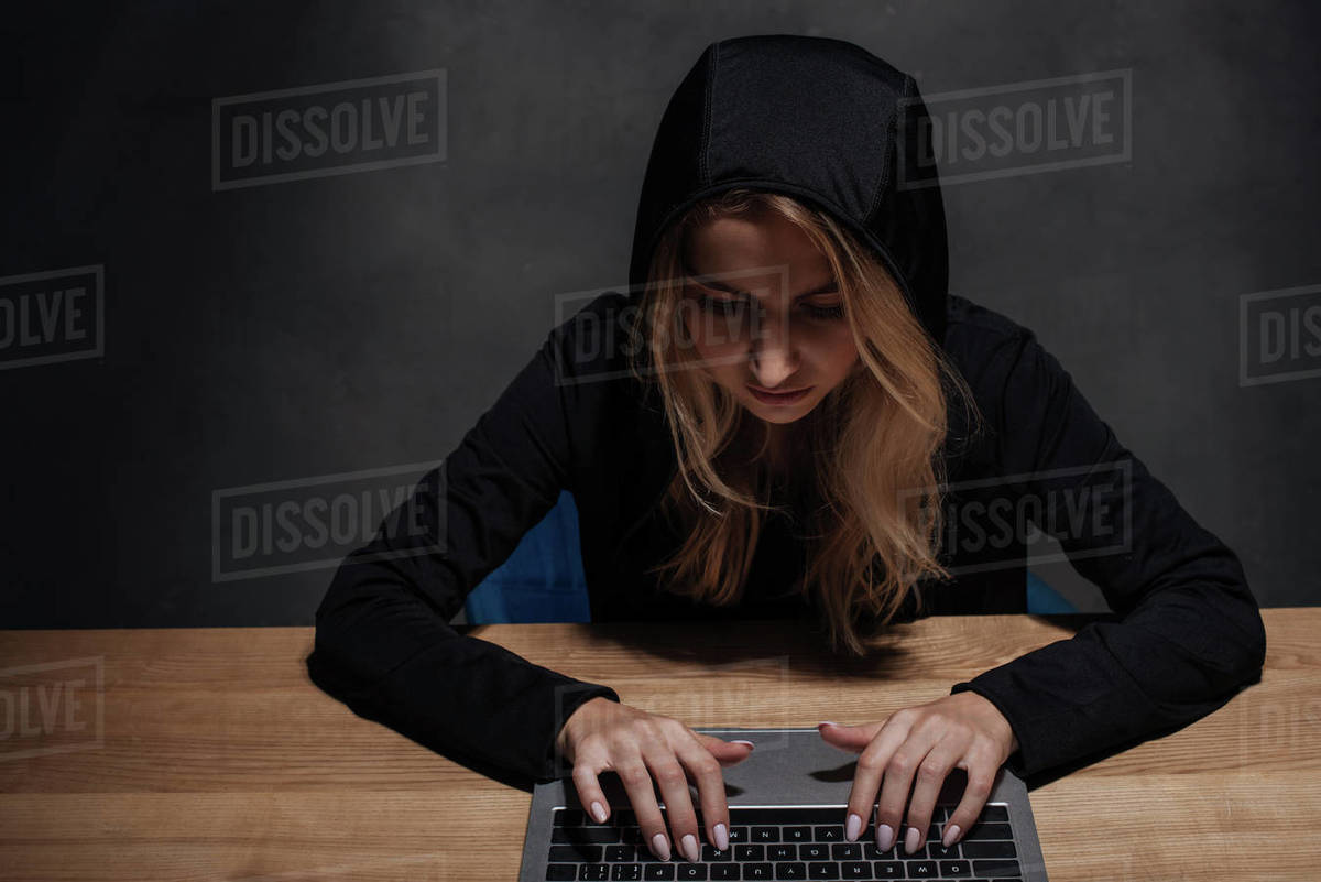 Focused female hacker in black hoodie using laptop at wooden tabletop ...