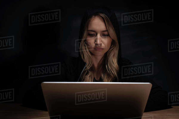 Portrait of female hacker in black hoodie using laptop at wooden ...