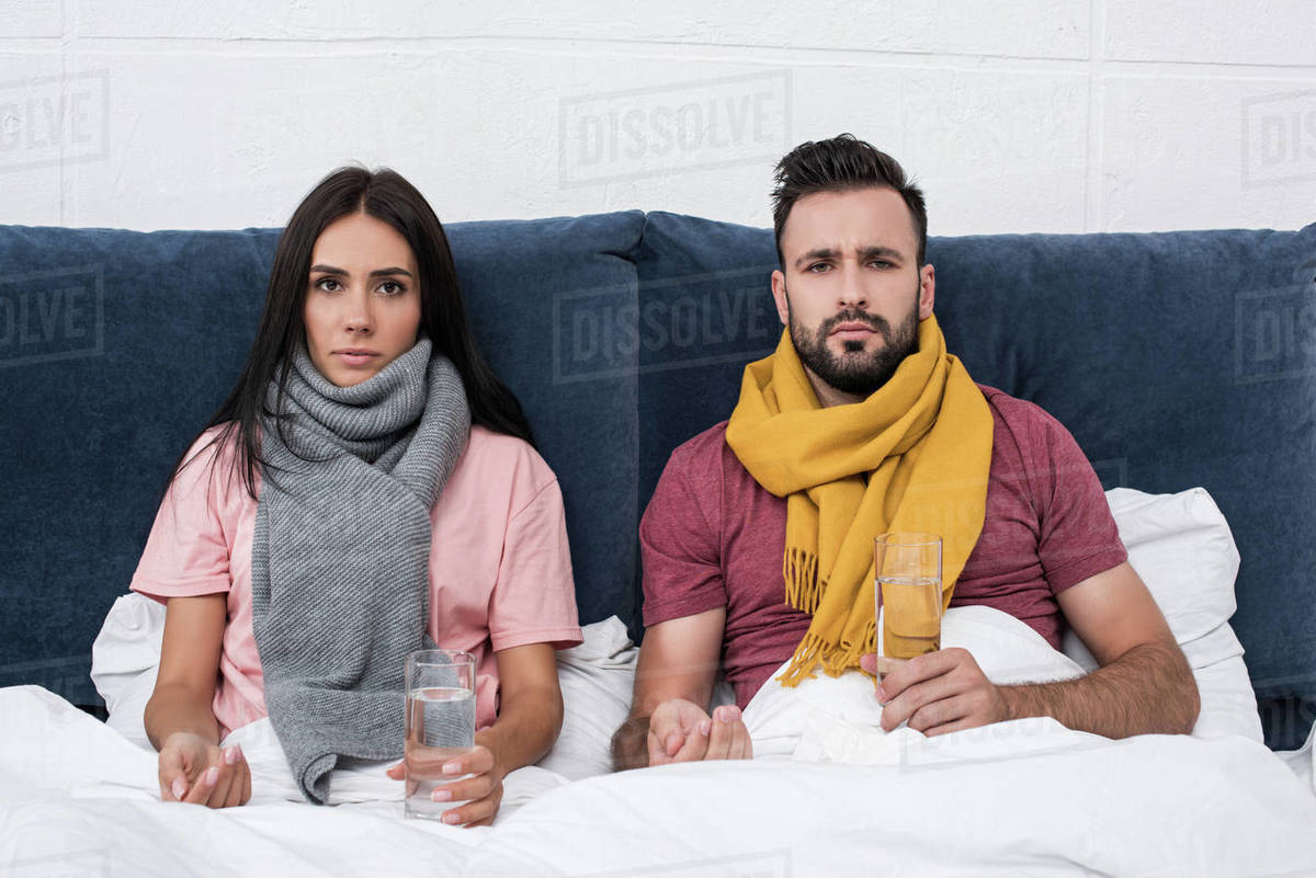 Serious sick couple with pills and glasses of water sitting in bed and ...
